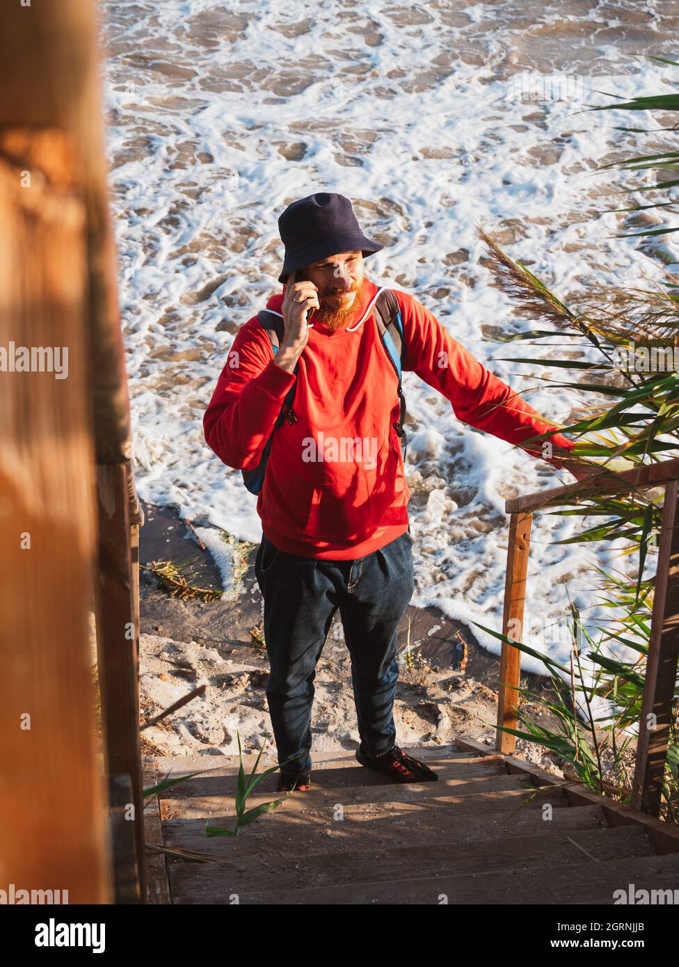 Bearded millennial man in red hoody with backpack autumn sea background ...