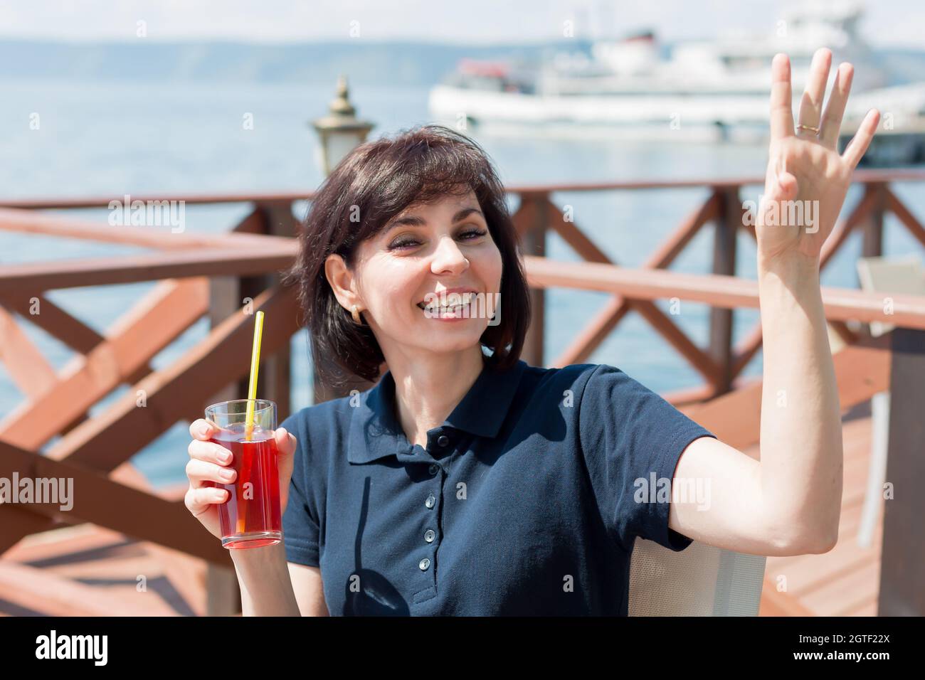 A brunette in a cafe by the sea with a glass of juice happily waves to ...