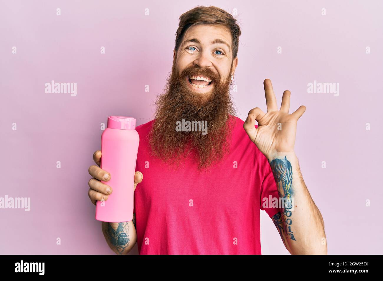 Redhead man with long beard holding shampoo bottle doing ok sign with ...