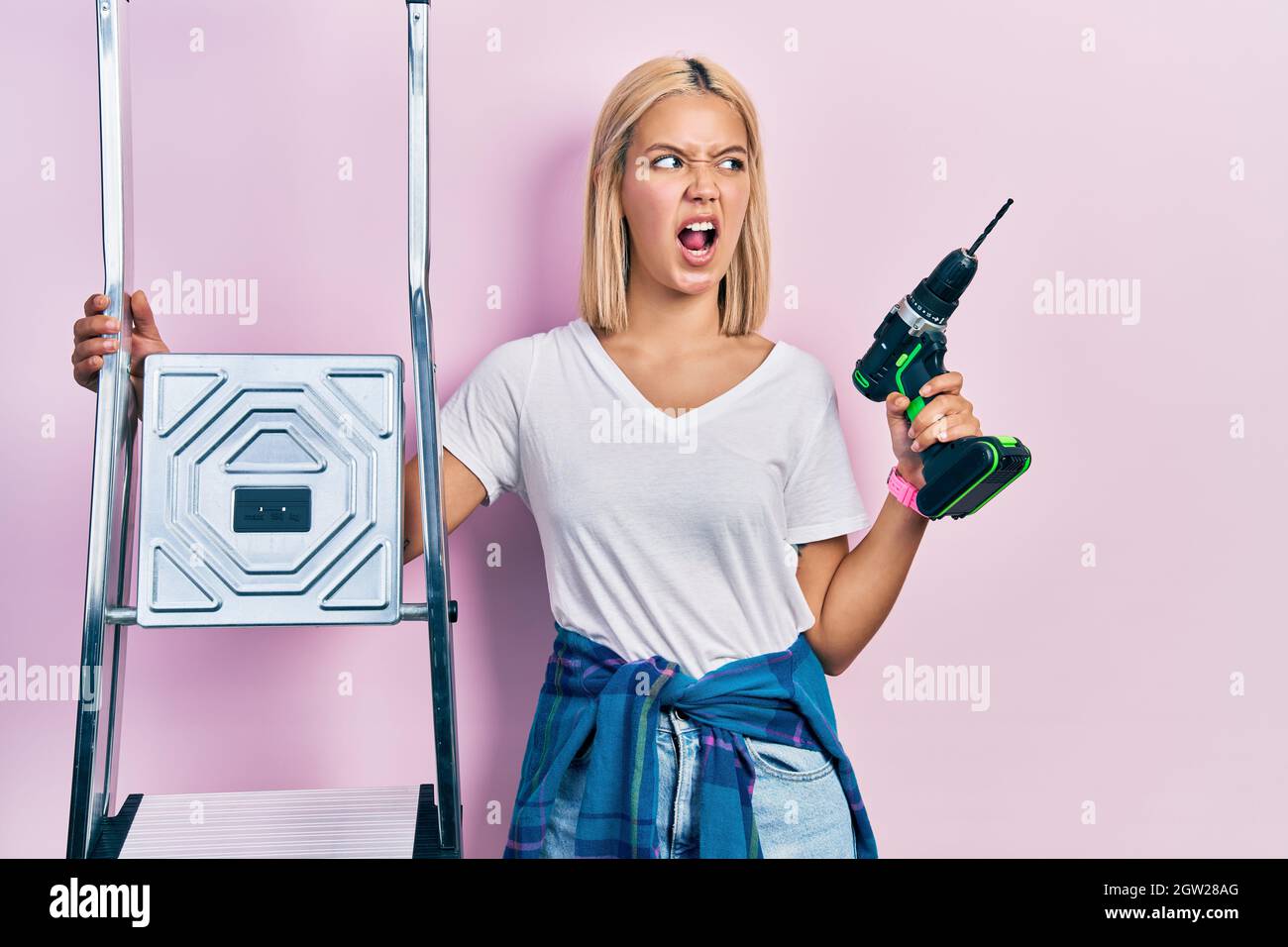 Beautiful blonde woman holding ladder holding screwdriver angry and mad ...