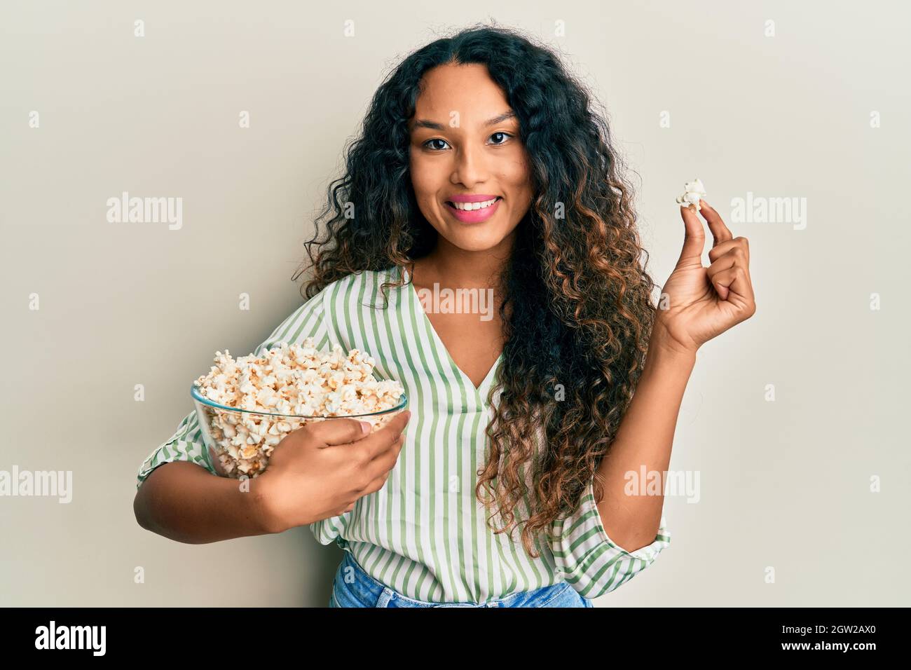 Young latin woman eating popcorn smiling with a happy and cool smile on ...
