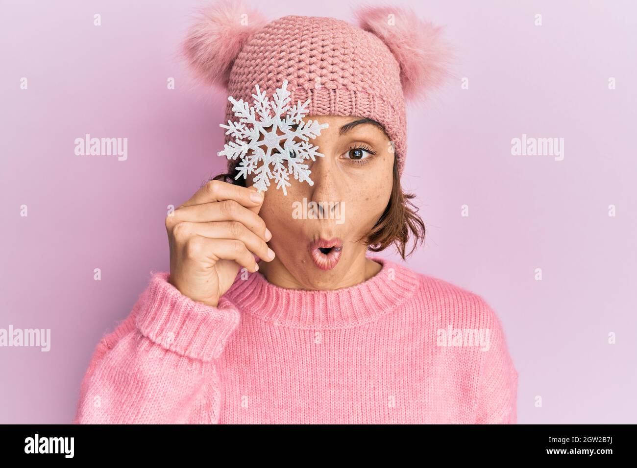 Young brunette woman holding snowflake wearing winter cap scared and ...