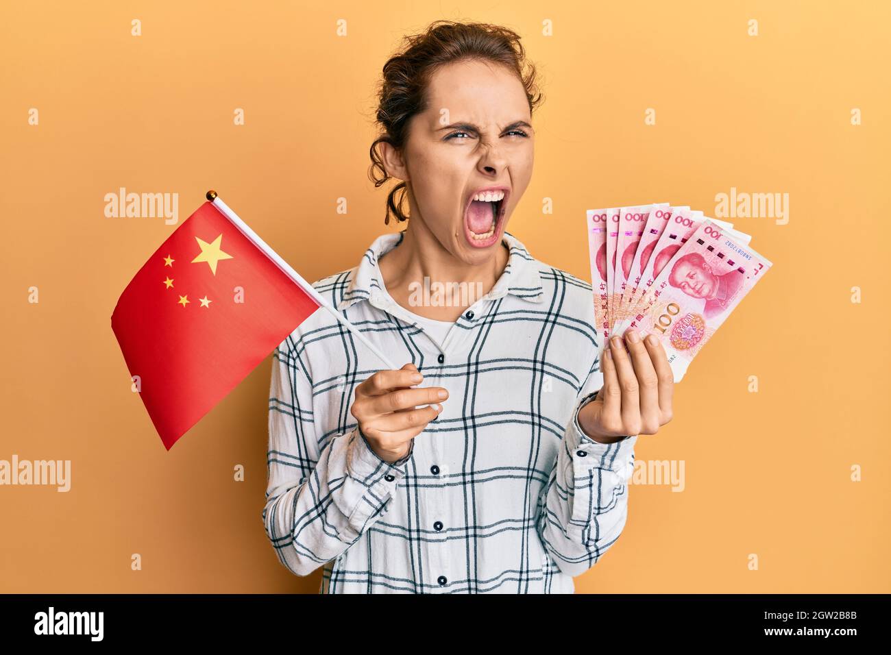 Young brunette woman holding china flag and yuan banknotes angry and ...