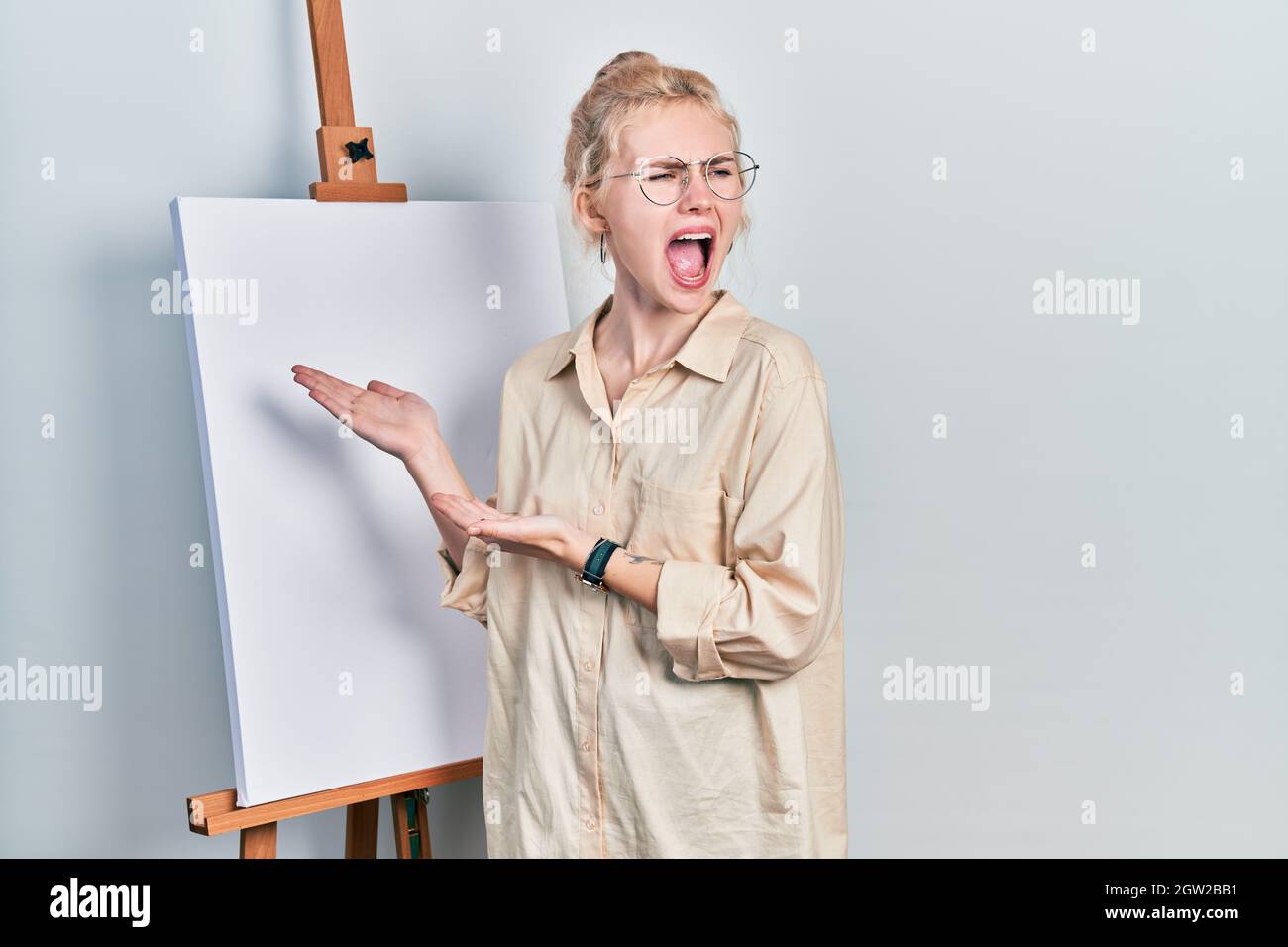 Beautiful caucasian woman with blond hair standing by painter easel ...