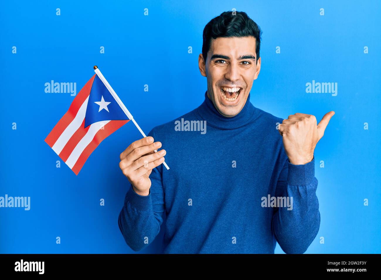 Handsome hispanic man holding puerto rico flag pointing thumb up to the ...
