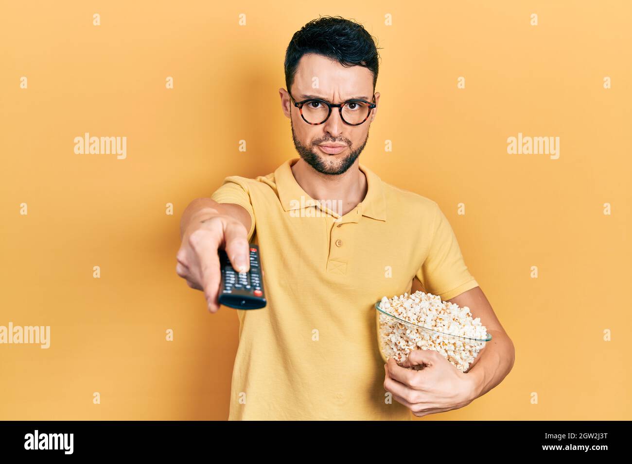 Young hispanic man eating popcorn using tv control skeptic and nervous ...