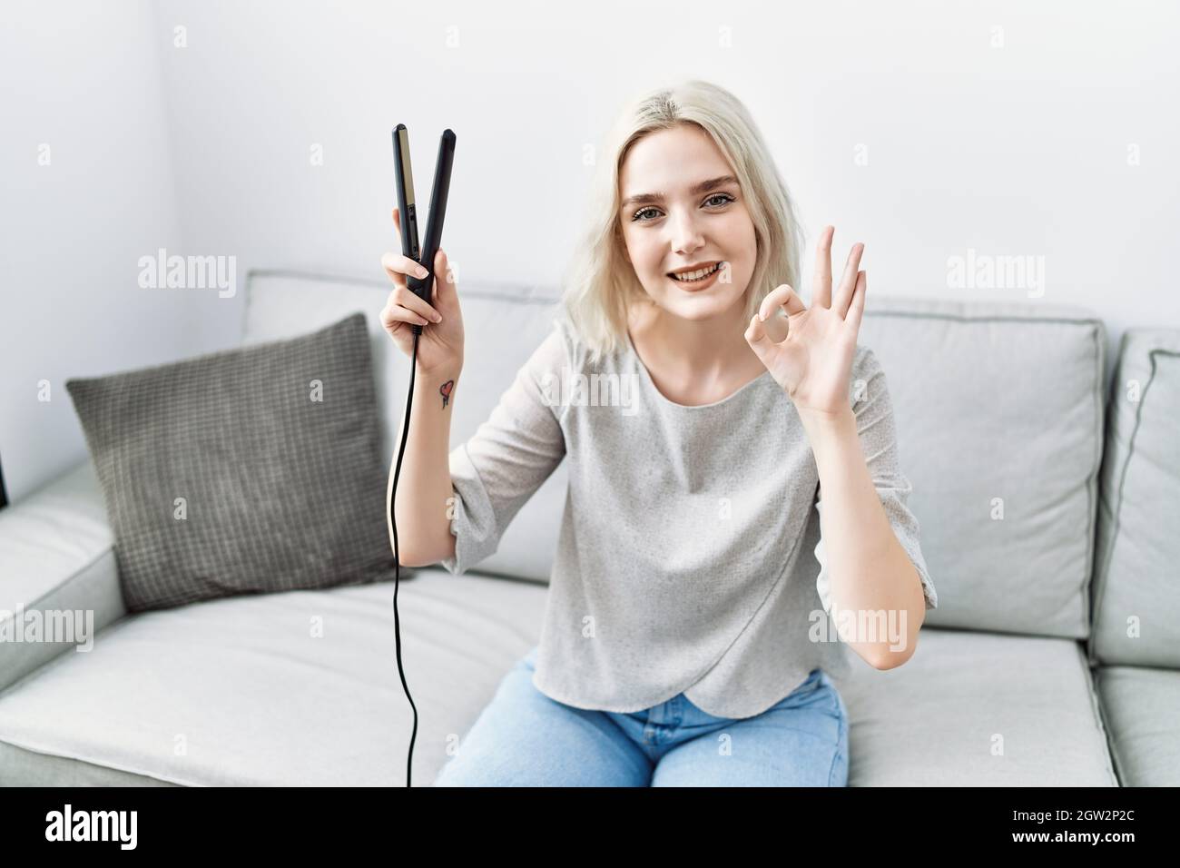 Young caucasian woman at home holding hair straightener doing ok sign ...