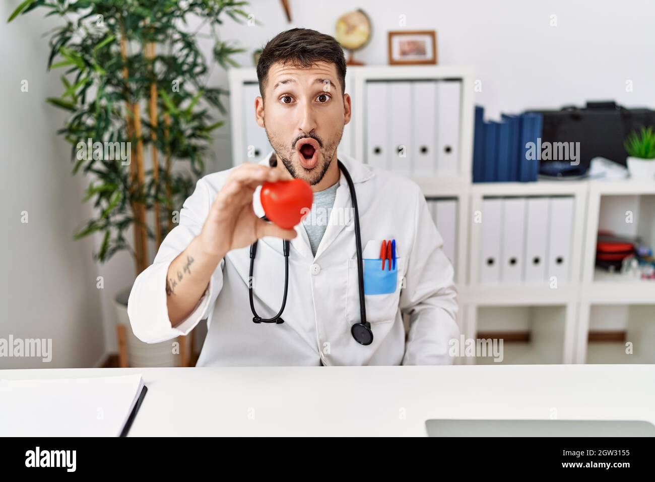 Young doctor holding heart at medical clinic scared and amazed with ...