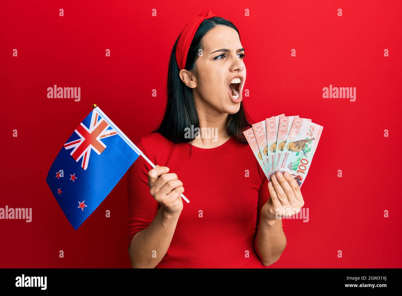 Young hispanic woman holding new zealand flag and dollars angry and mad ...