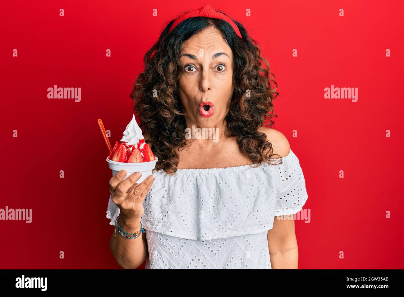 Middle age hispanic woman eating strawberry ice cream scared and amazed ...