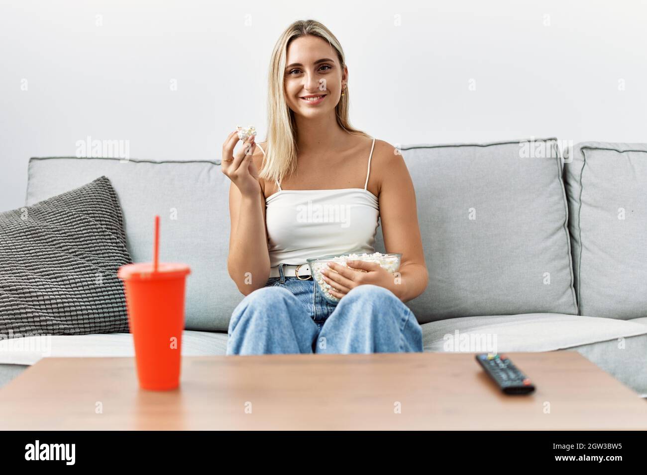 Young blonde woman eating popcorn sitting on the sofa looking positive ...