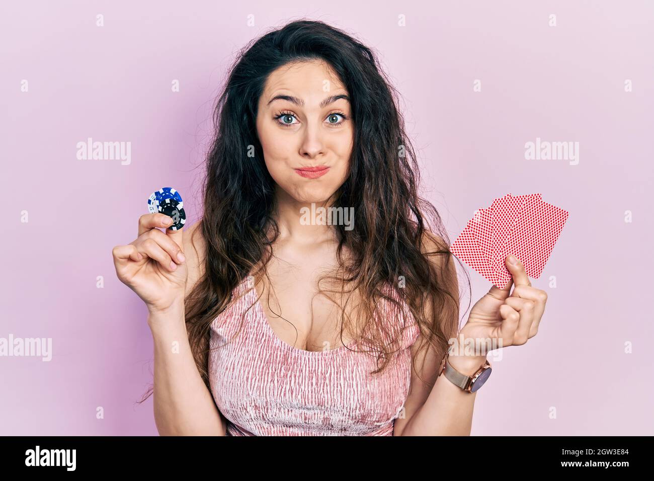 Young hispanic woman playing poker holding casino chips and cards puffing cheeks with funny face ...