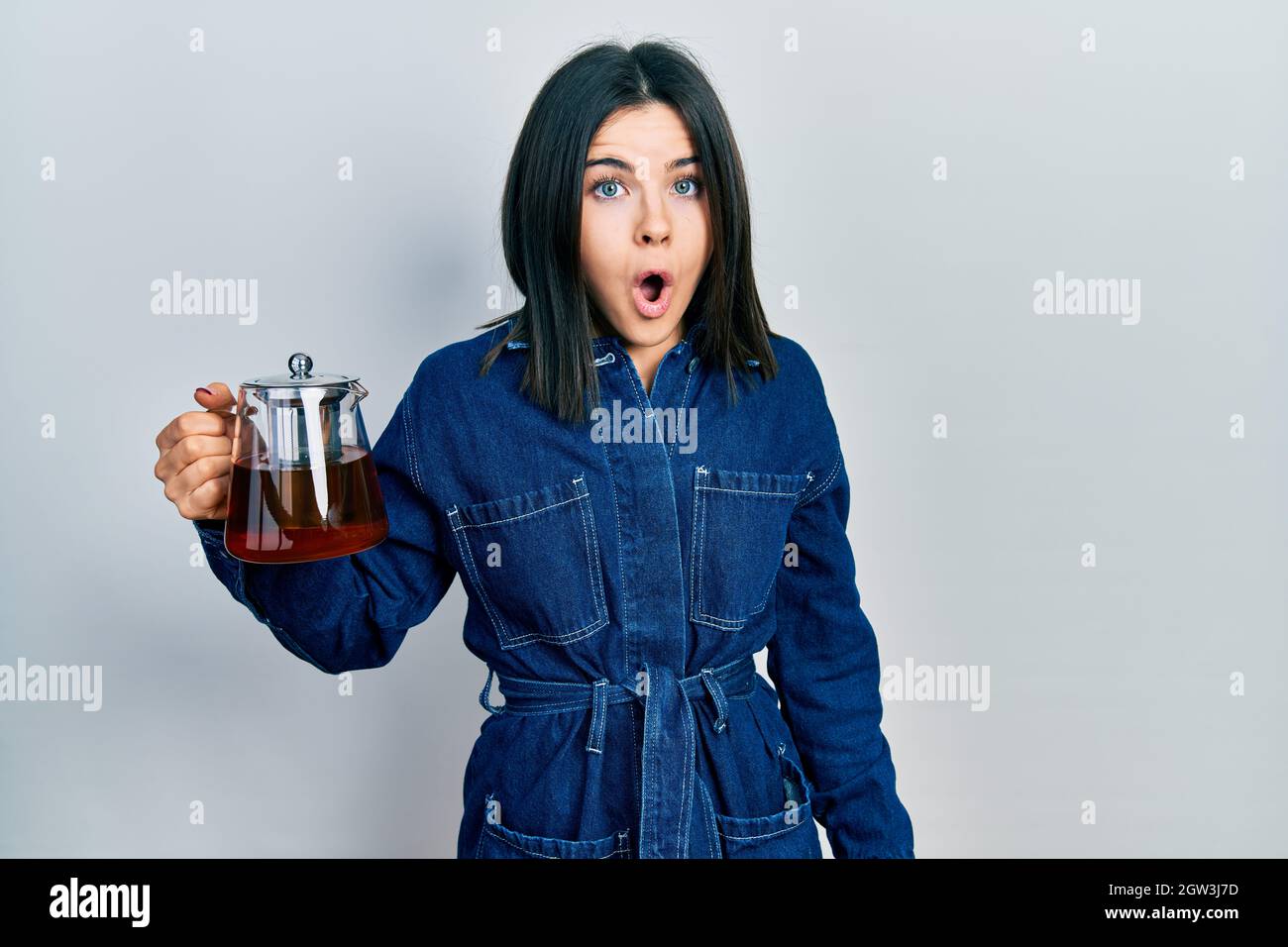 Young brunette woman wearing traditional tea pot scared and amazed with ...