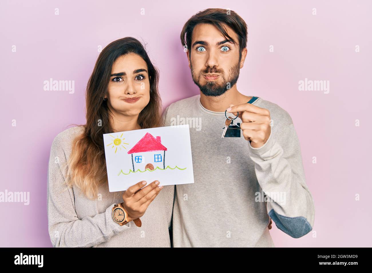 Young hispanic couple holding house draw and key of new home puffing cheeks with funny face ...