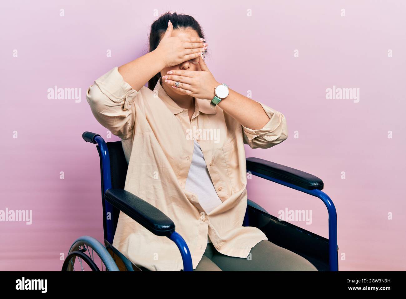 Young hispanic woman sitting on wheelchair covering eyes and mouth with ...