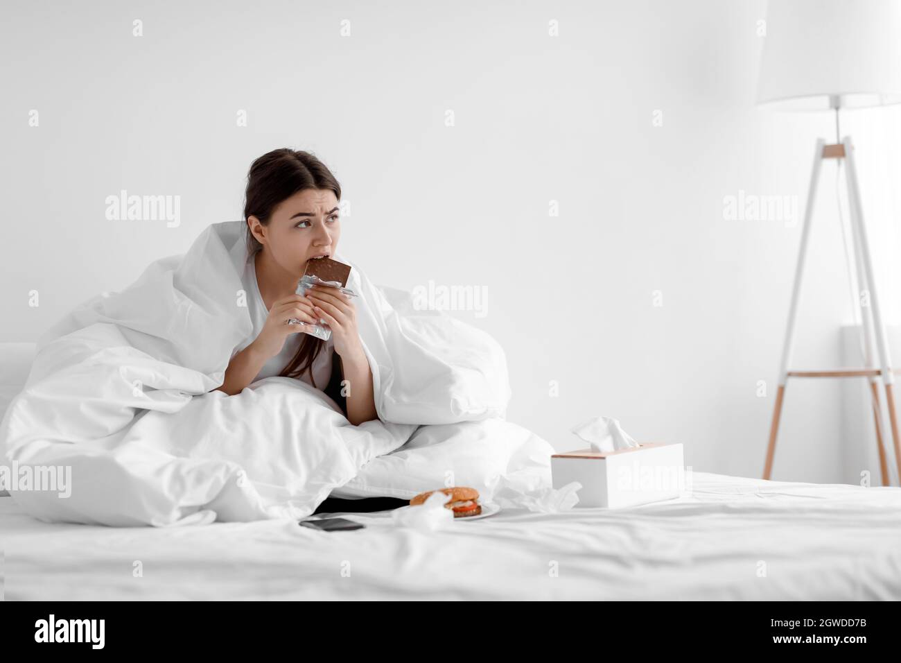 Upset hungry caucasian young woman sitting in bed at home and eating ...