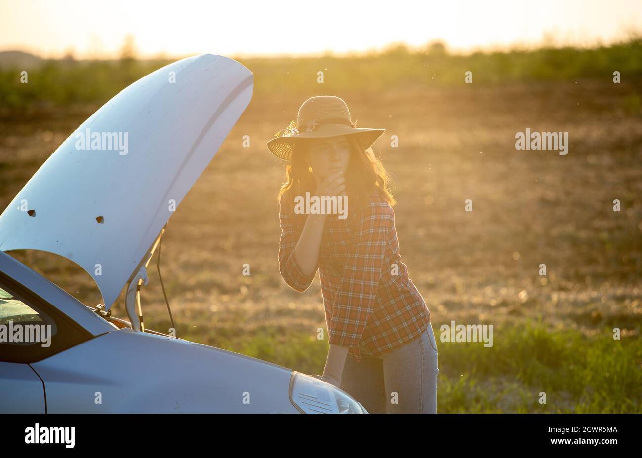 Young woman standing in front of car with popped hood thinking ...