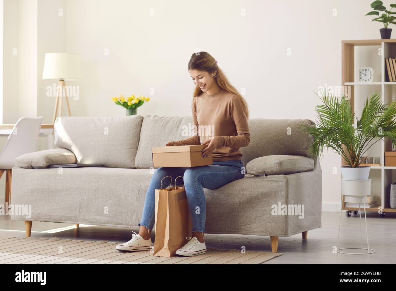 Happy young woman sitting on sofa at home unpacks cardboard parcel with ...