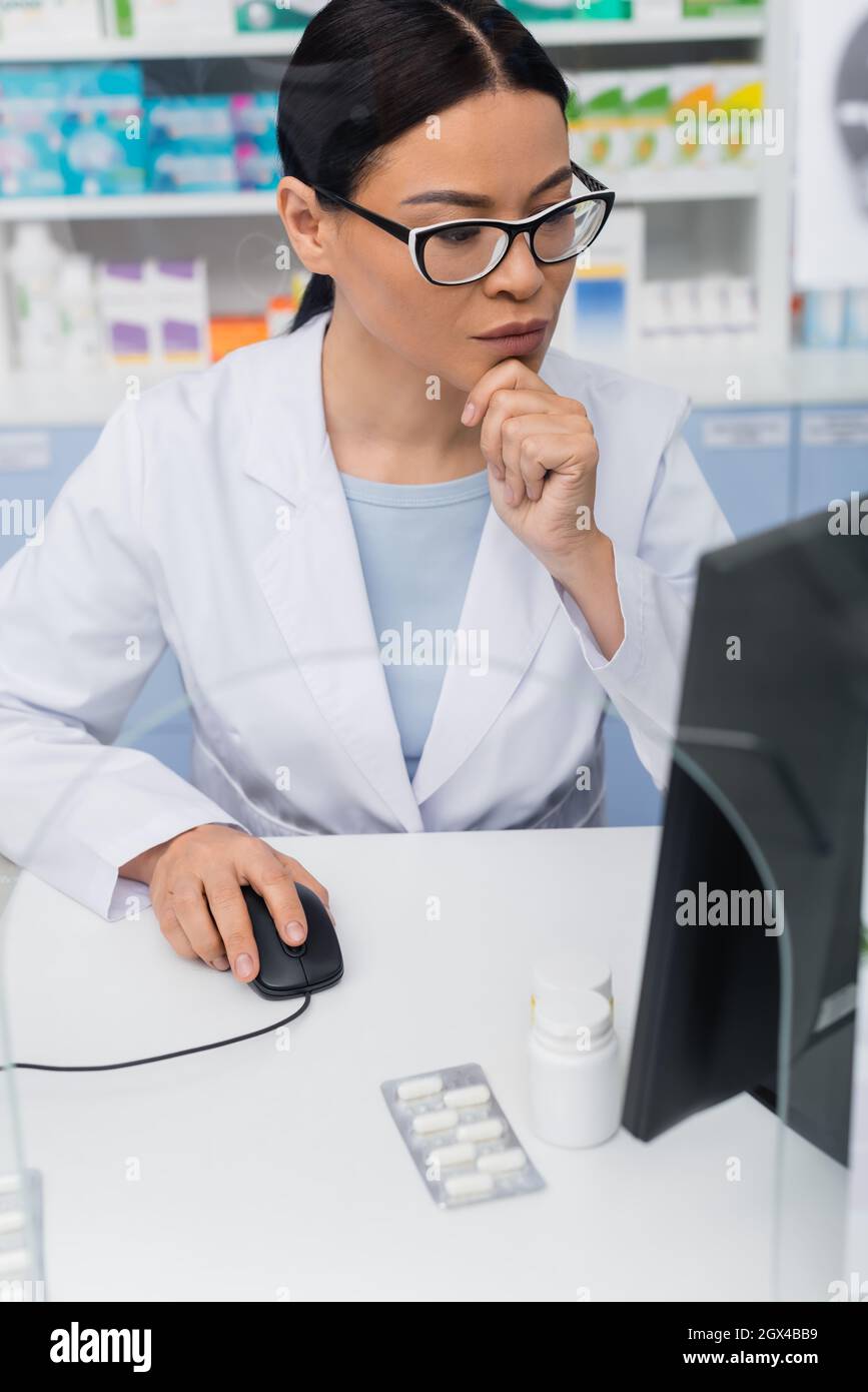 brunette asian pharmacist in glasses and white coat using computer in ...