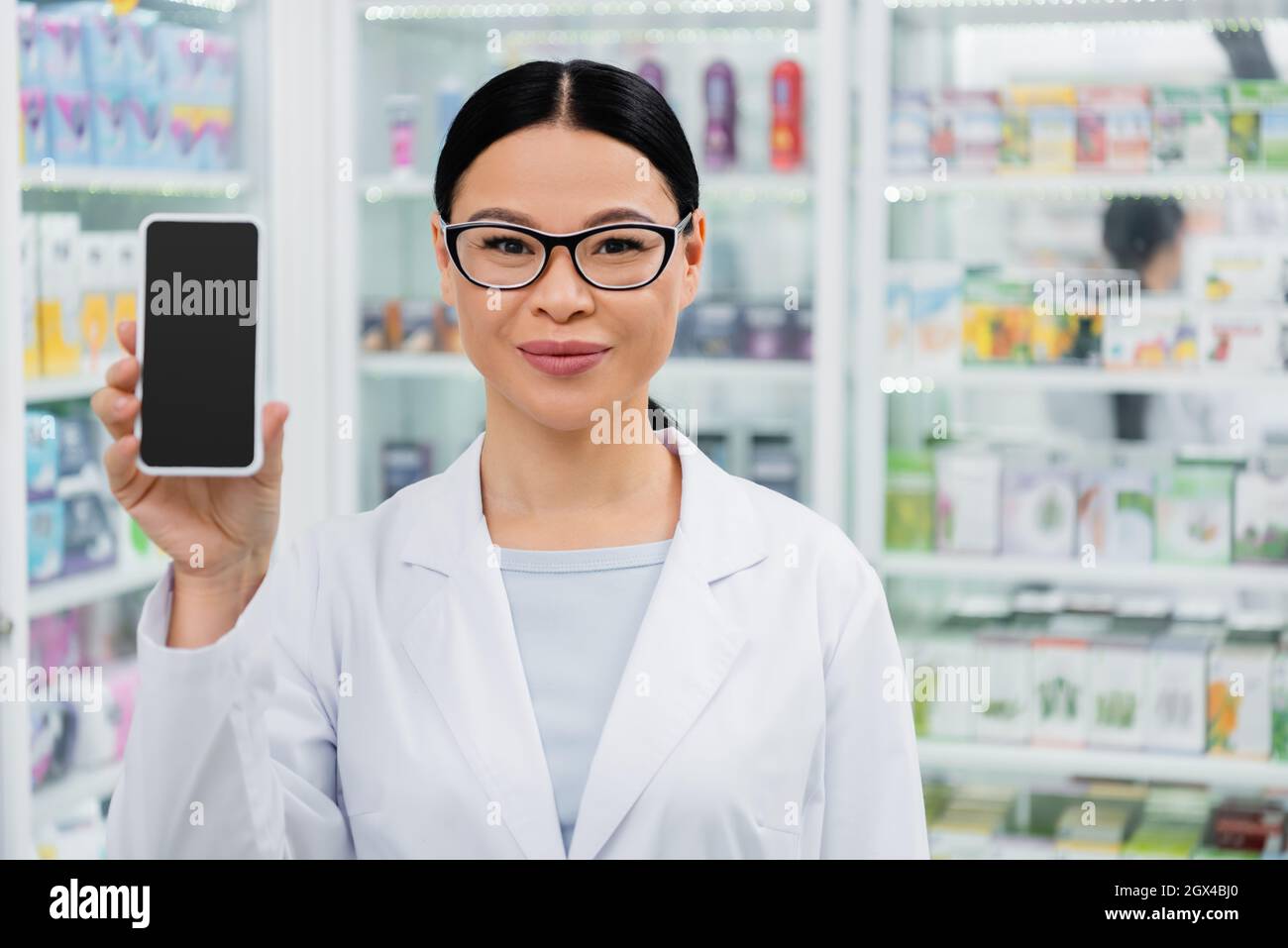 asian pharmacist in glasses smiling while holding cellphone with blank ...