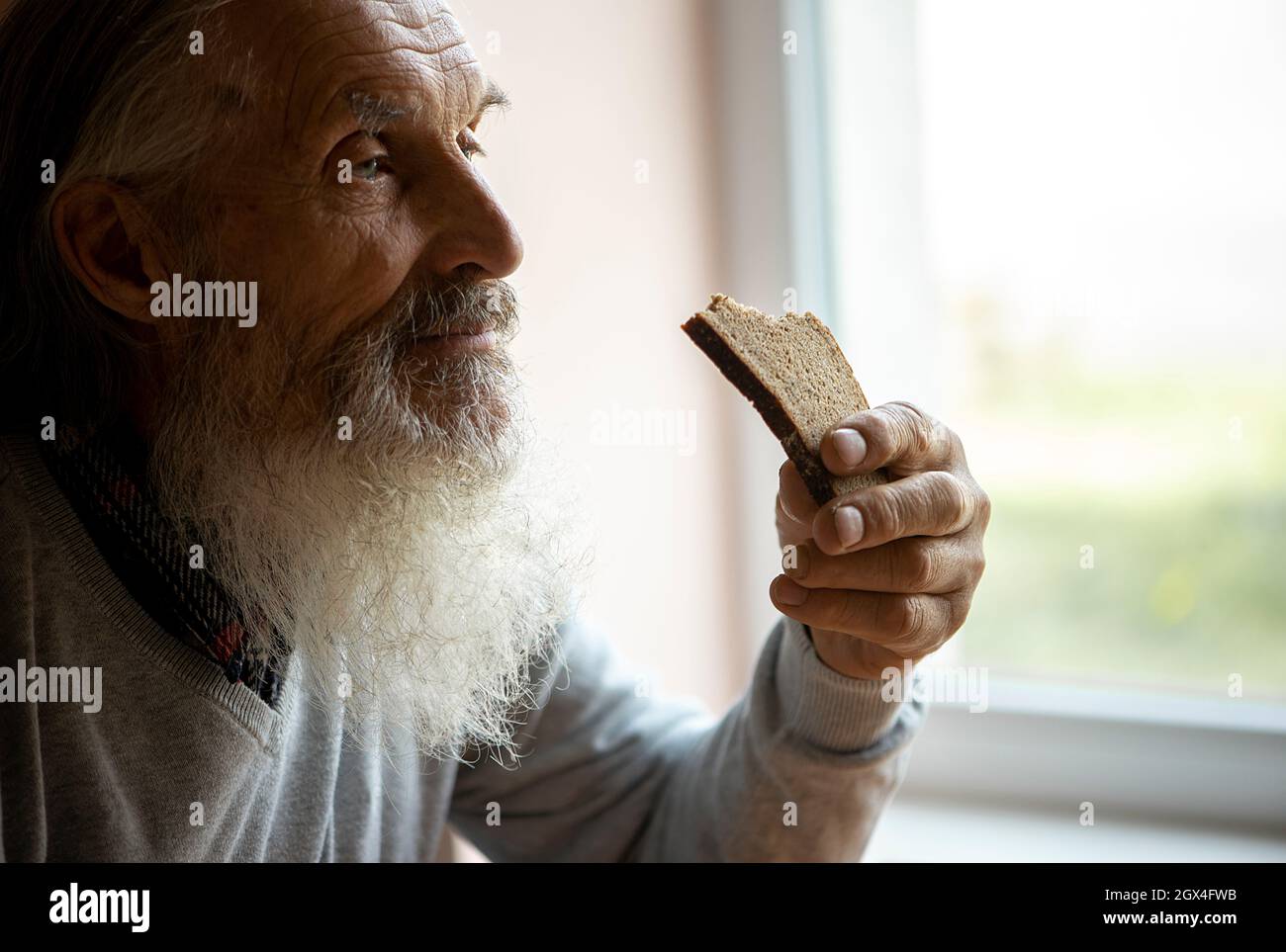 Old sad man with a long gray beard sitting by the window and eating ...
