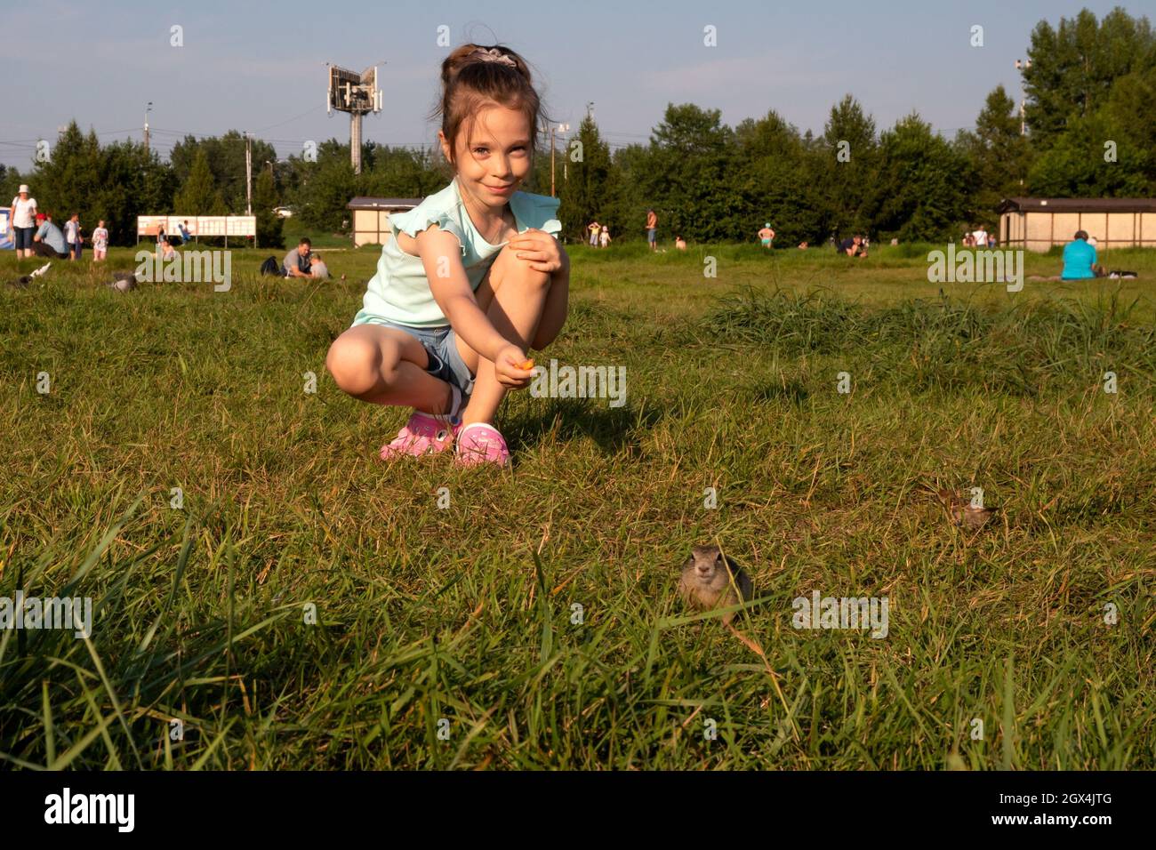 A smiling girl holds out a carrot to a gopher, in a meadow with people ...