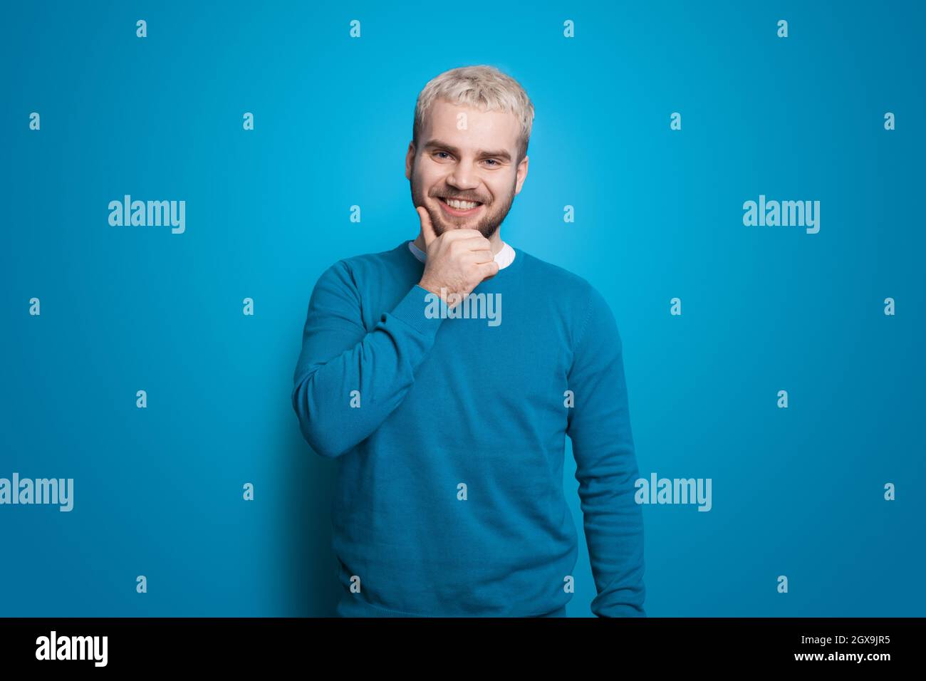 Man with blonde hair on blue wall is touching chin. Smiling man. Front ...