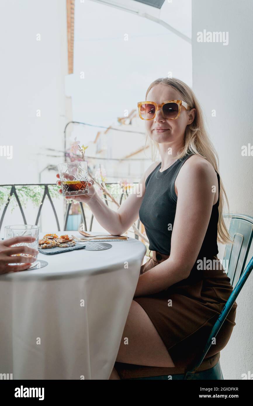 Side view of young female in sunglasses sitting with glass of whiskey ...