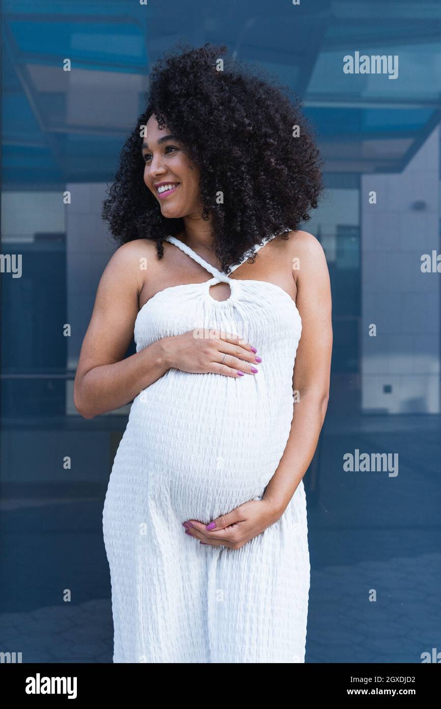 Charming pregnant African American with Afro curls wearing white dress standing near reflecting ...