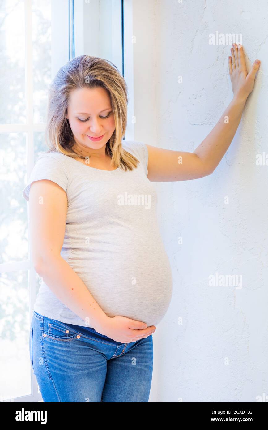 Beautiful pregnant woman standing near window at home in light interior ...