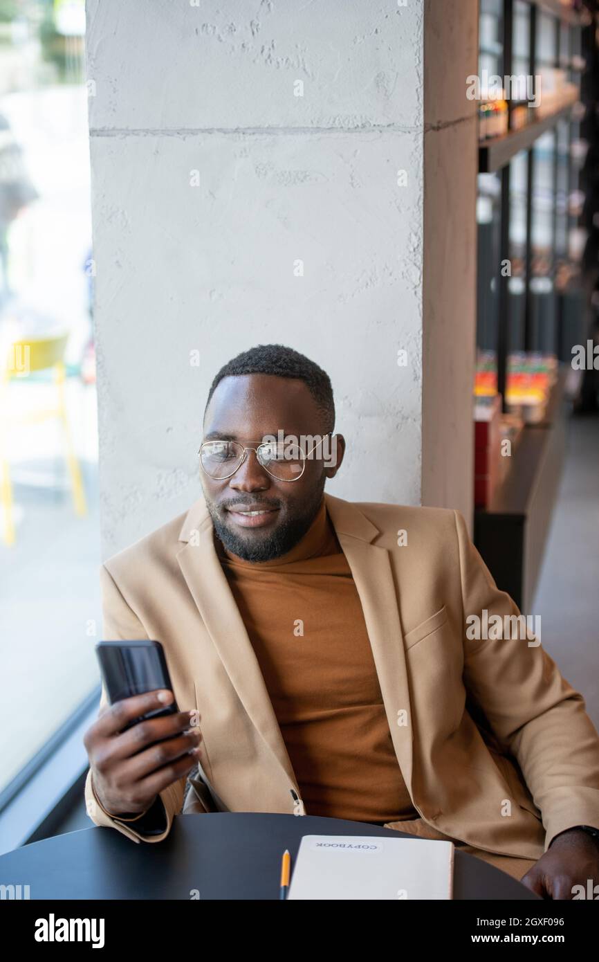 Young elegant African man scrolling in smartphone by table in bookshop ...