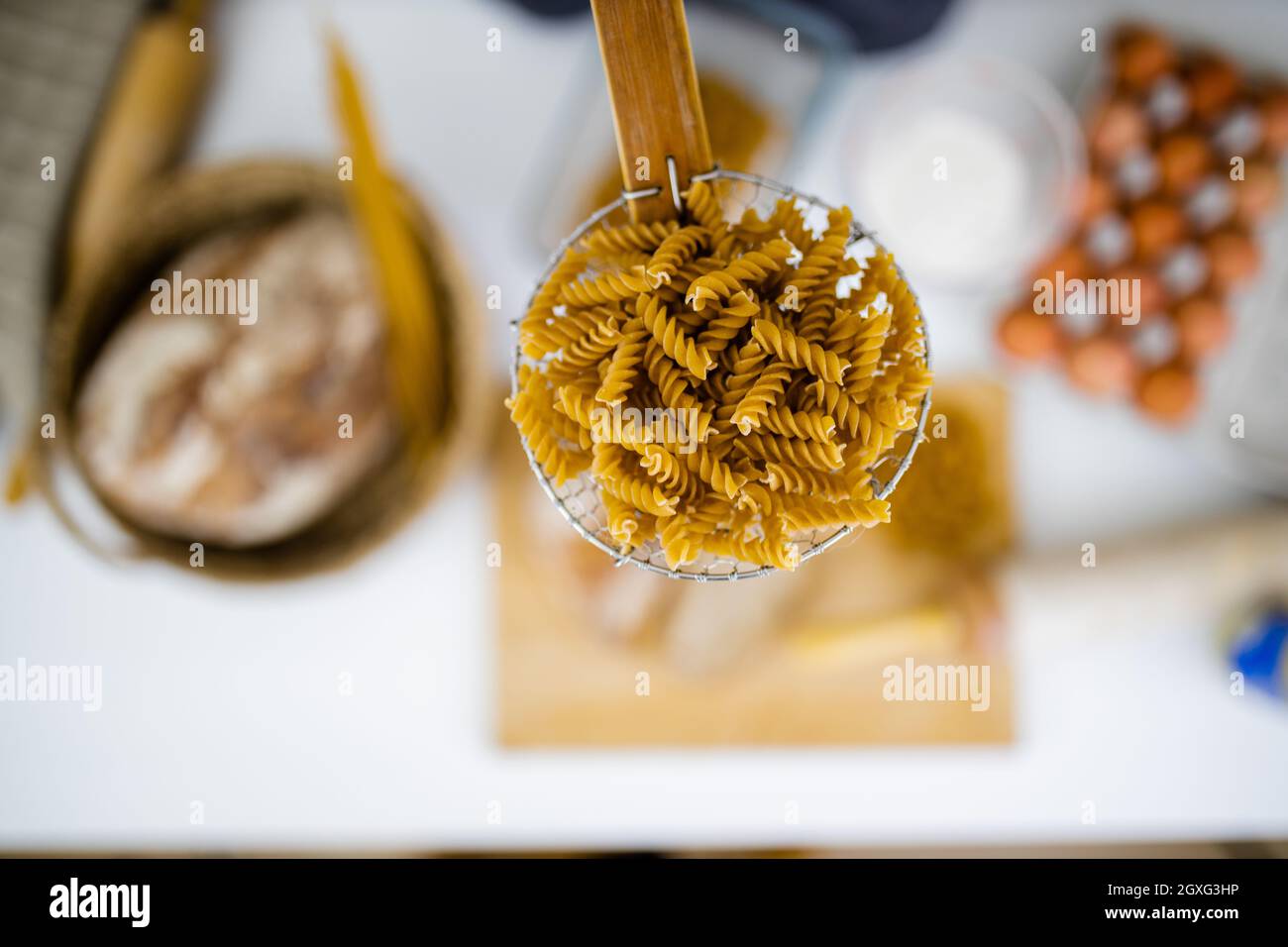 Uncooked fusilli on wooden pasta strainer above blurry ingredients on ...