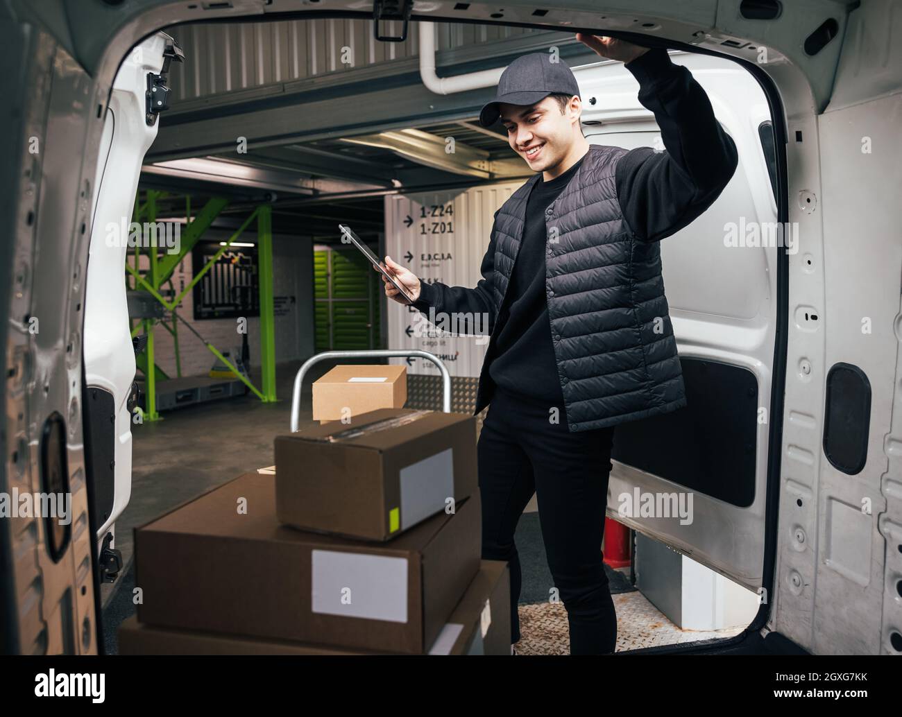 Smiling courier standing at the trunk of a van checks cardboard boxes ...