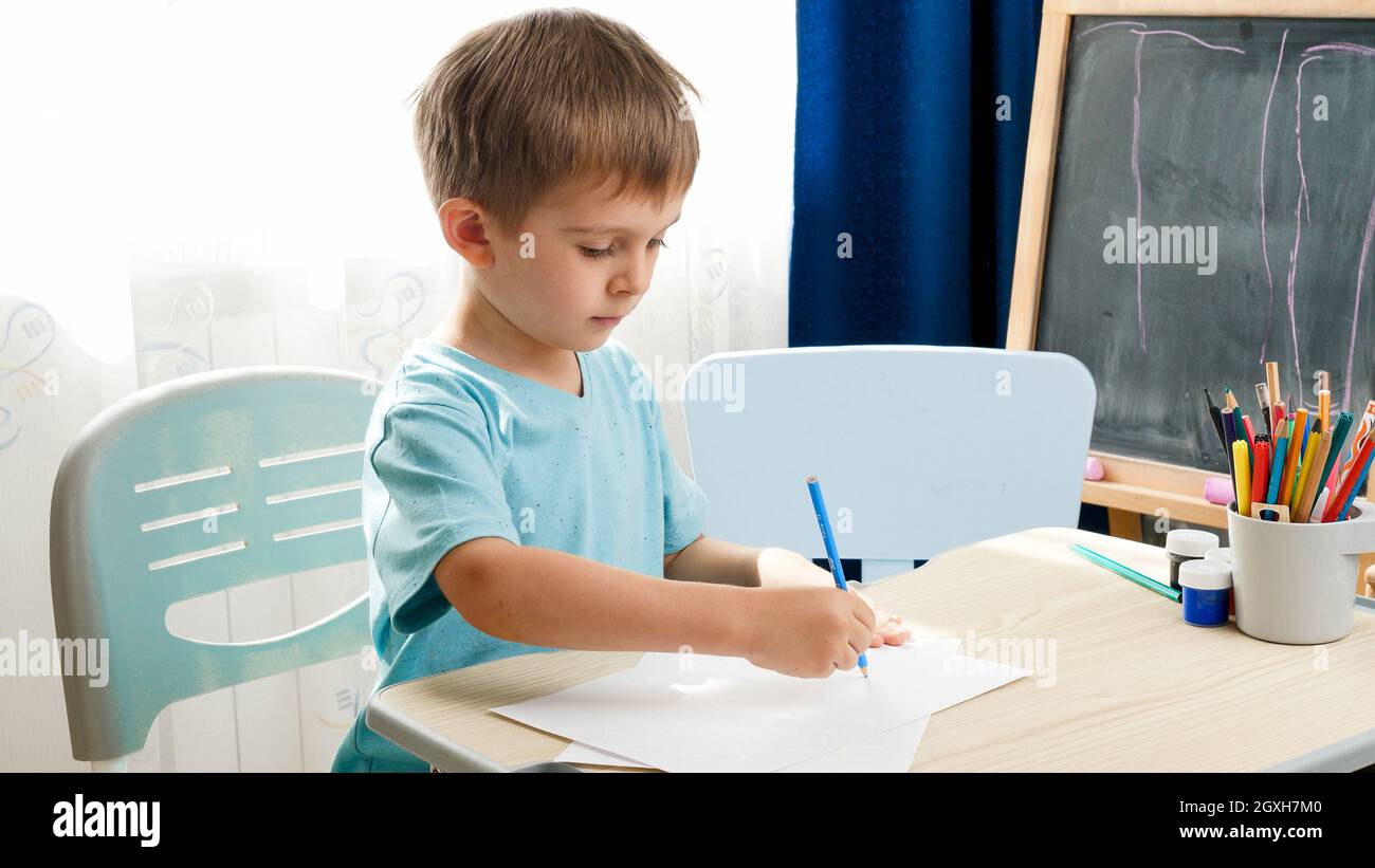 Concentrated little boy drawing or writing with pencil while sitting ...