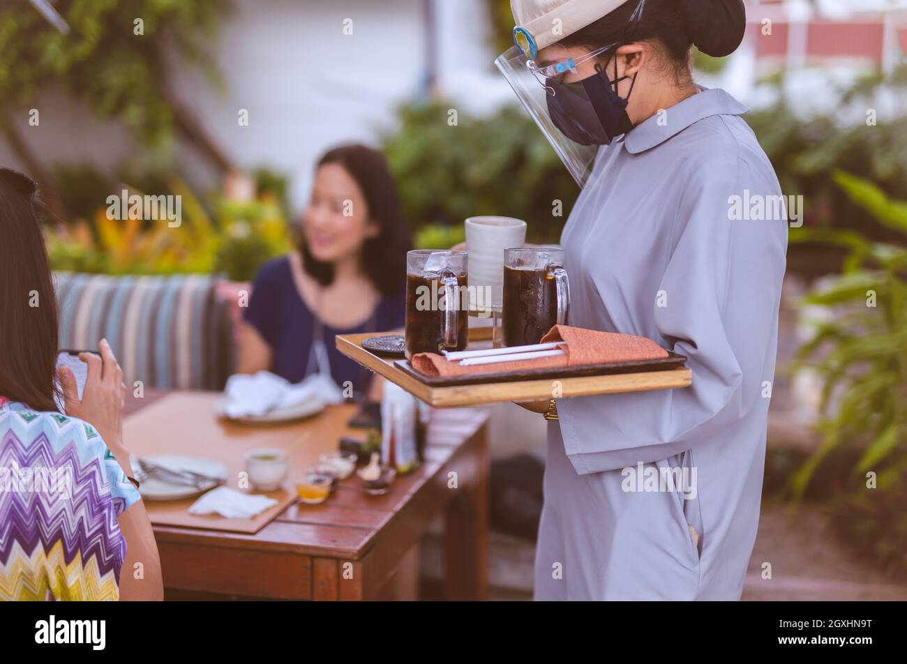 Female waiterss in face shield and mask serves iced coffee to customer ...