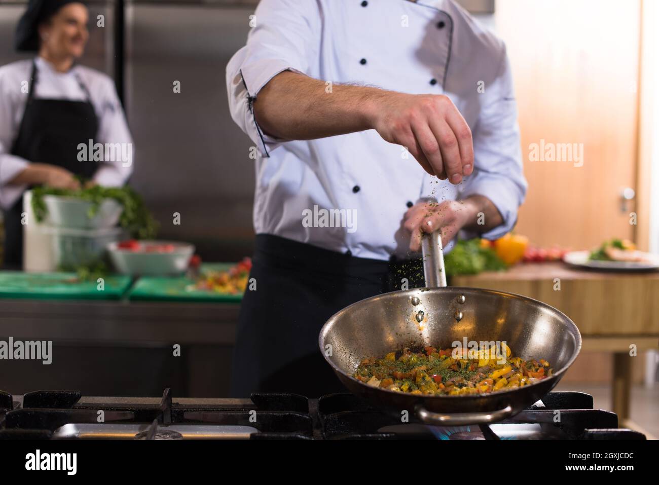 Young male chef putting spices on vegetables in wok at commercial ...
