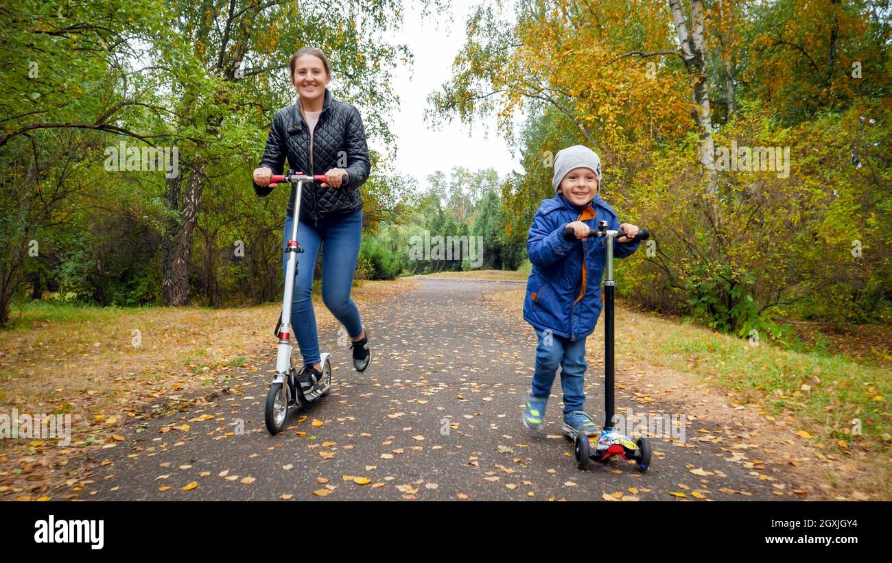 Happy smiling boy with mother racing on kick scooters at autumn park ...