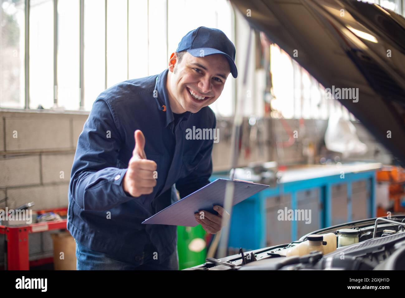Smiling mechanic giving thumbs up in front of a car with the hood open ...