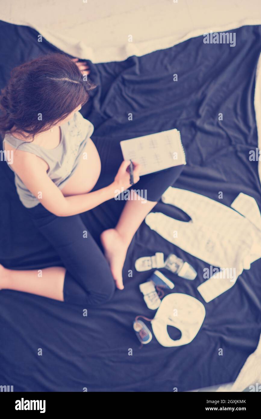 a pregnant woman at home on the floor checking list of baby clothes ...