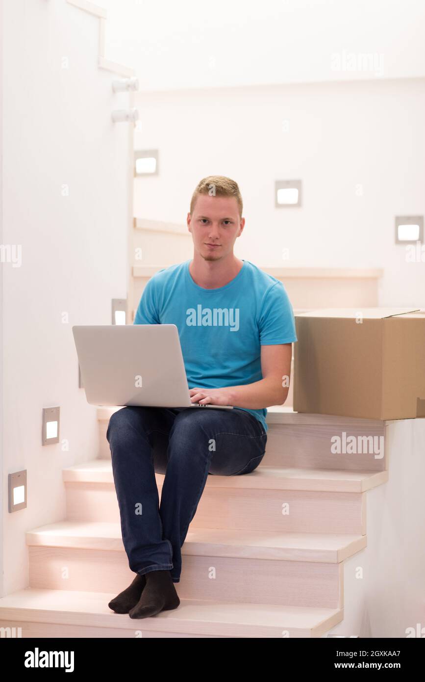Happy young man sitting in stairway at home, using laptop computer with ...
