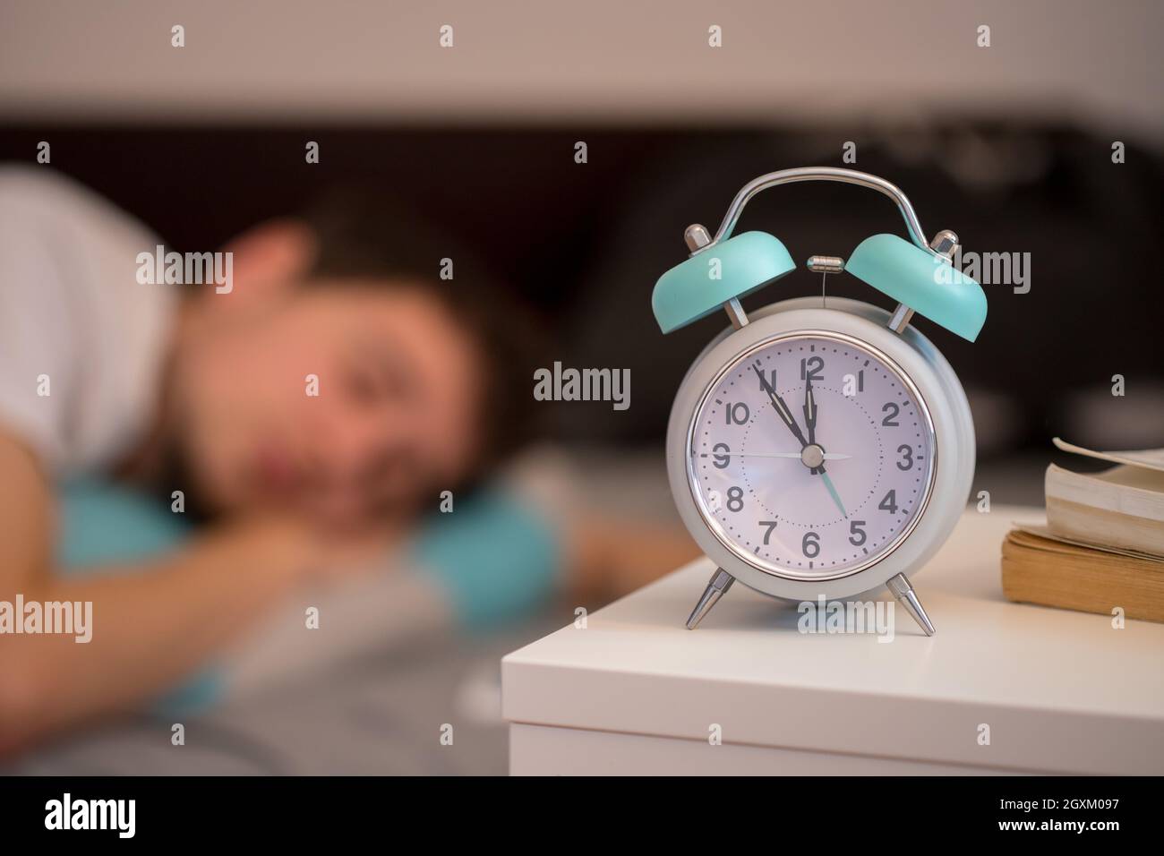 White alarm clock on the bedside table. Young man sleeps in the ...