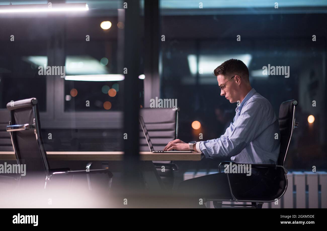 Young man working on laptop at night in dark office. The designer works ...