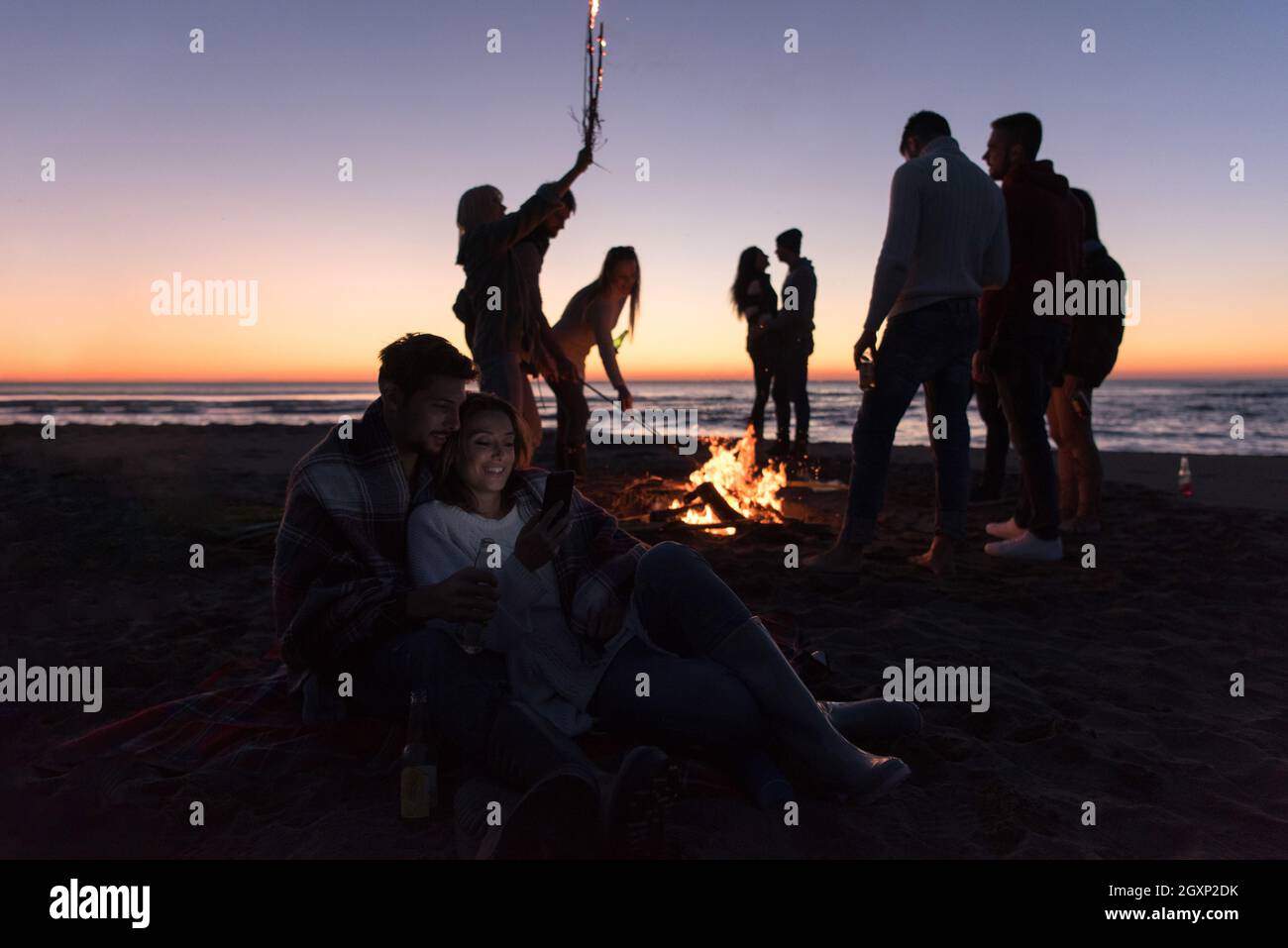 Couple using cell phone during autumn beach party with friends drinking ...
