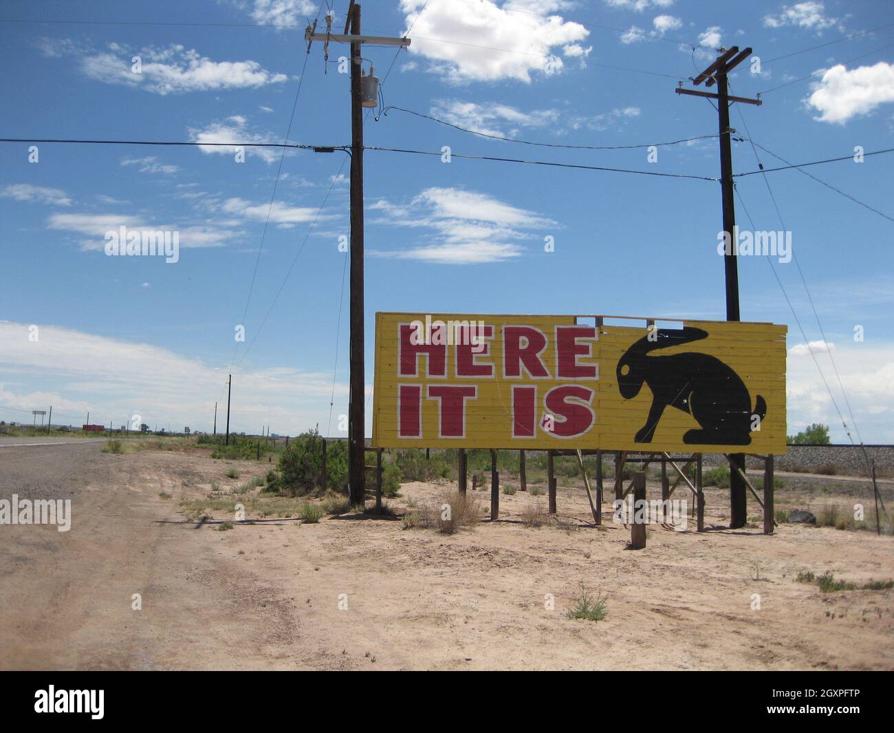 Along a desert road, a yellow billboard sign reads Here It Is with a ...