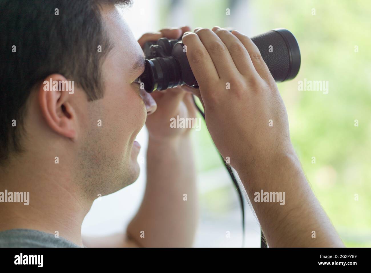 Young man standing looking through a glass window with binoculars as he ...