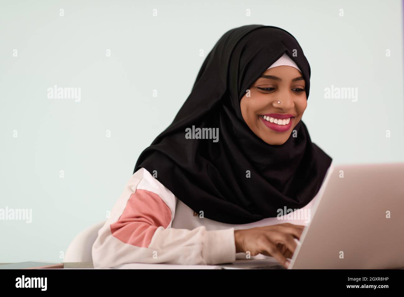 an afro muslim woman wearing a hijab sits smiling in her home office ...