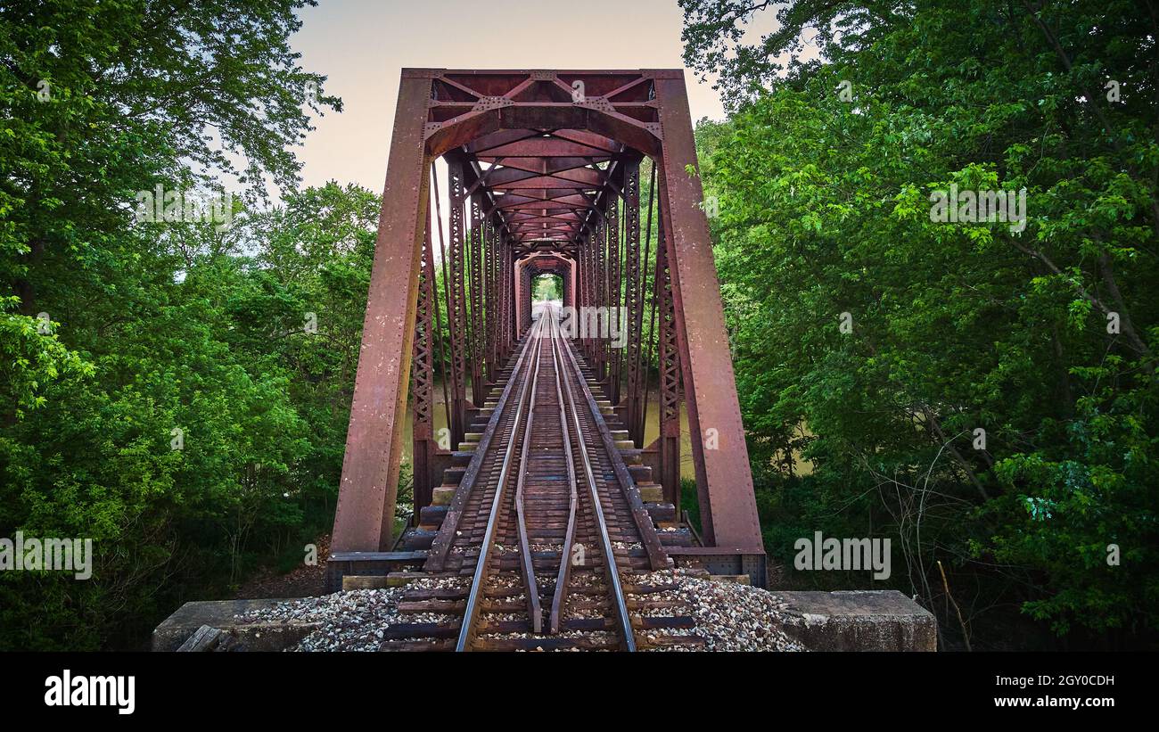 View straight on train tracks of large metal bridge over river in green ...