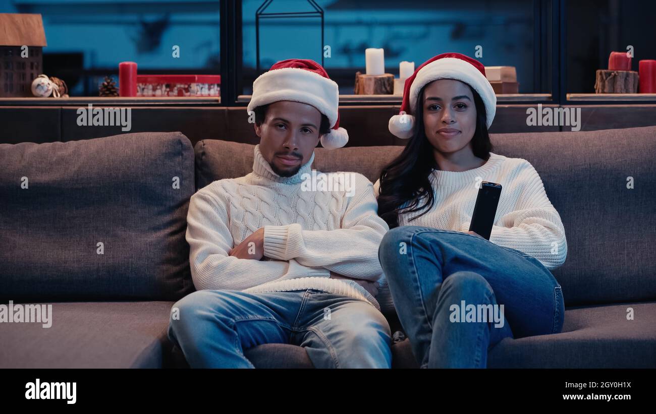 smiling african american woman watching movie with bored man in santa ...