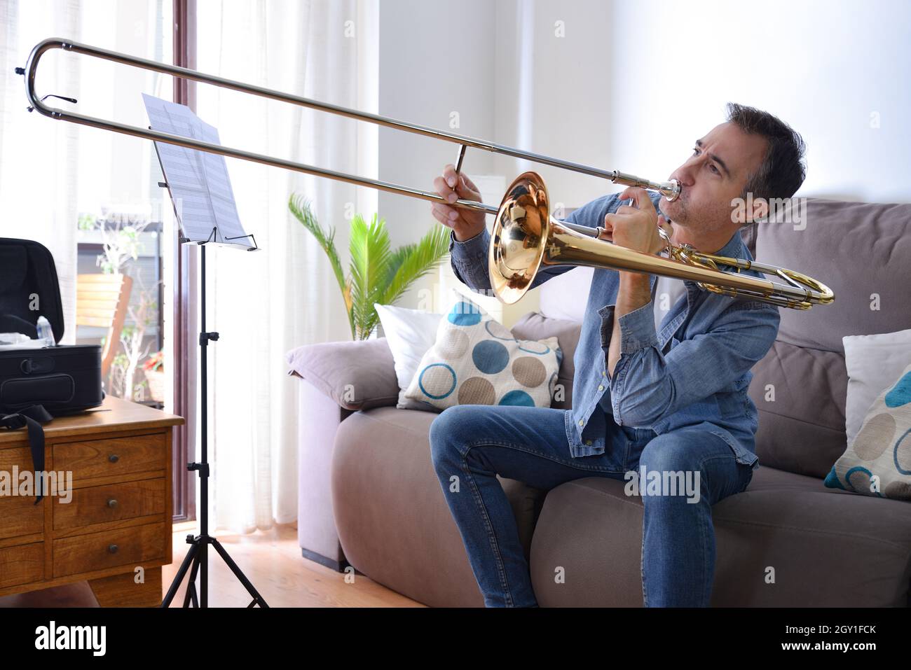 Novice man playing the trombone at home sitting on the sofa. Front view ...