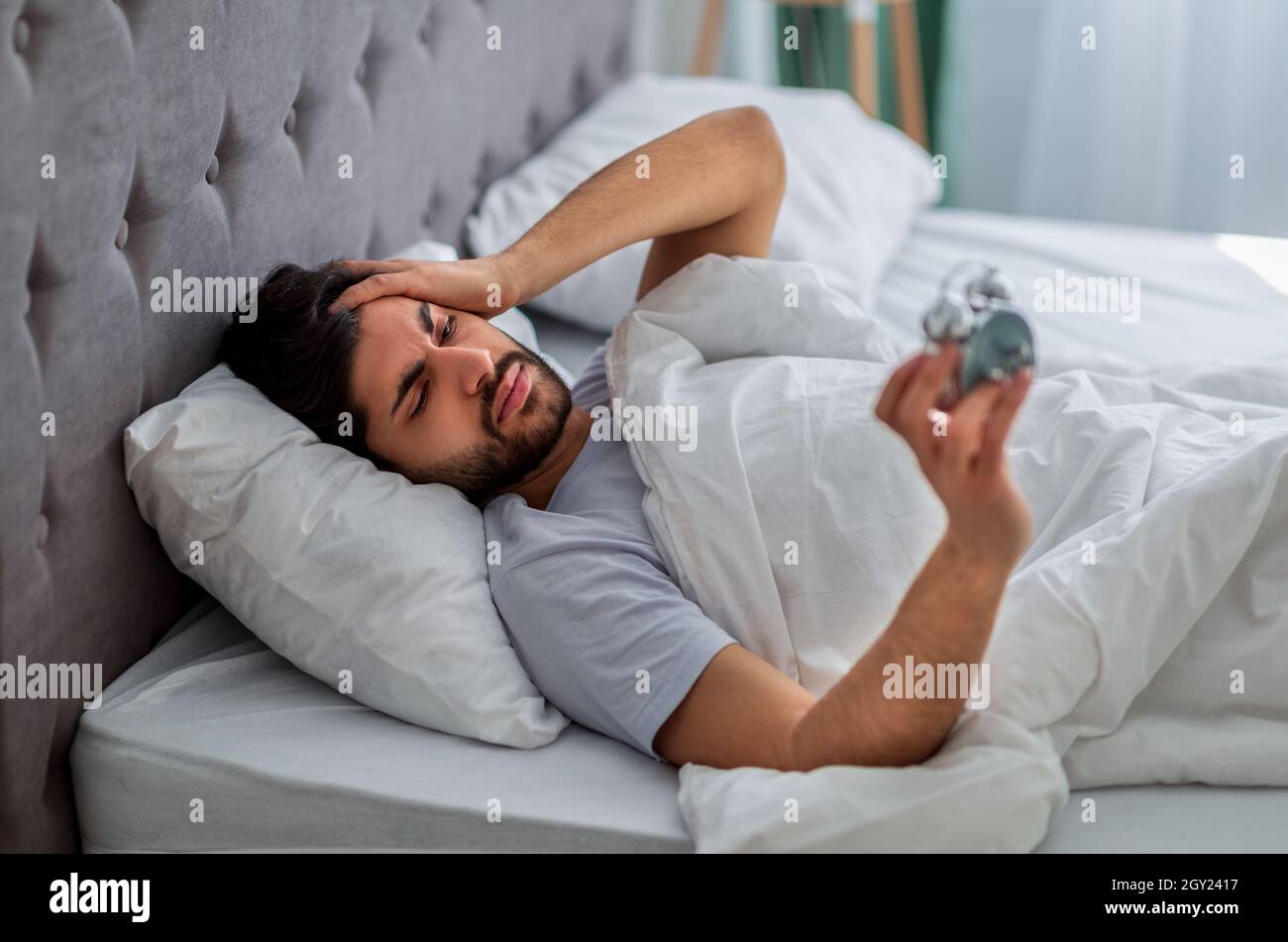 Unhappy arab man lying in bed with alarm clock, not willing to get up ...